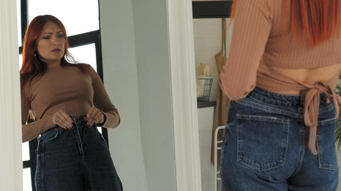 Upset redhead mature woman trying to zip tight jeans in front of